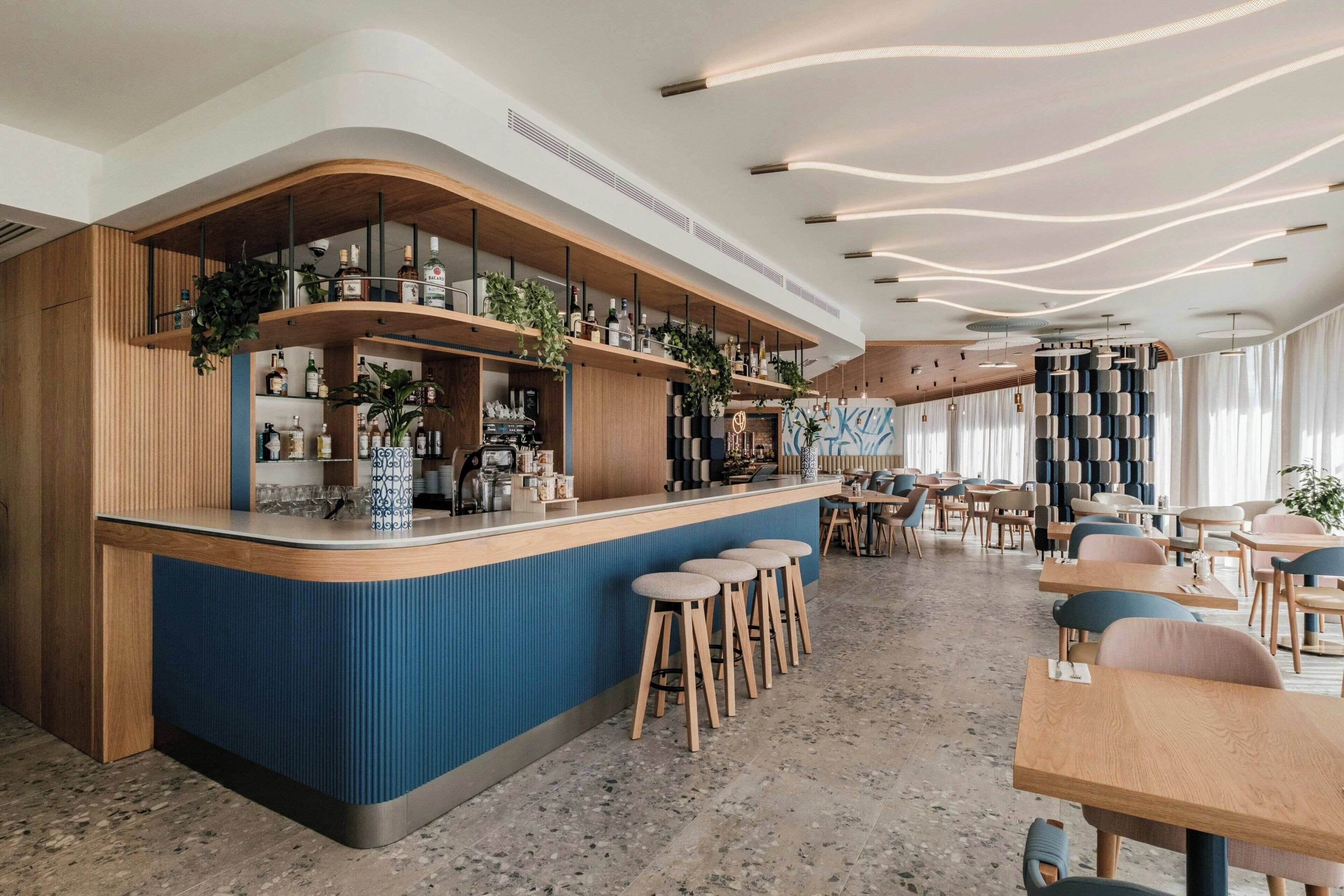 Uma área de bar moderna com balcão curvo, banquetas e diversos frascos e plantas.