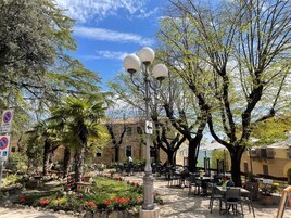 Um café ao ar livre com mesas e cadeiras, rodeado por árvores e um edifício ao fundo.