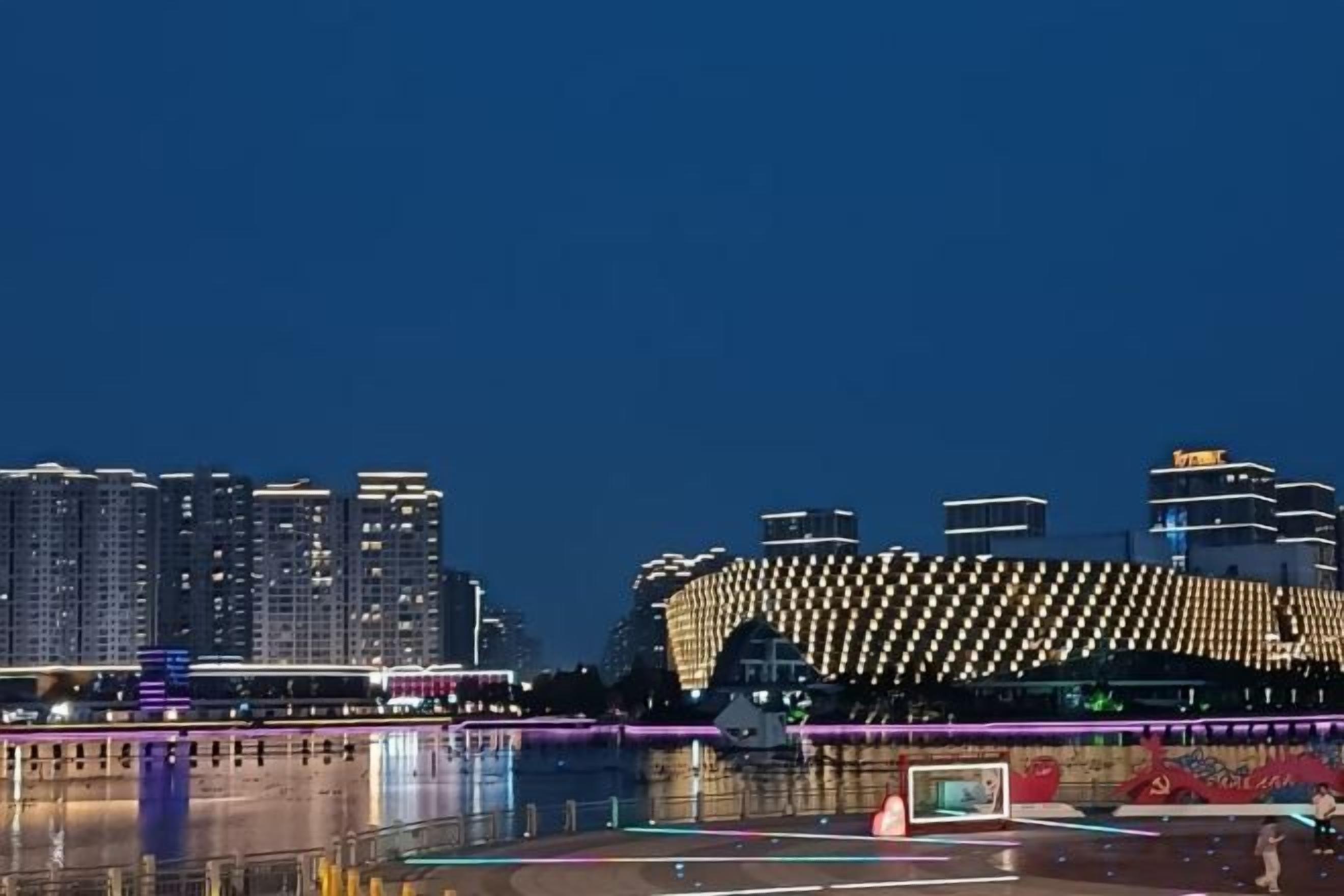 Paisagem urbana noturna com edifícios iluminados e uma estrutura moderna de grandes dimensões com um design de telhado singular.