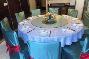 Mesa de jantar redonda com tampo de vidro, rodeada por cadeiras cor verde-água com fitas vermelhas, disposta com pratos, tigelas e talheres brancos.