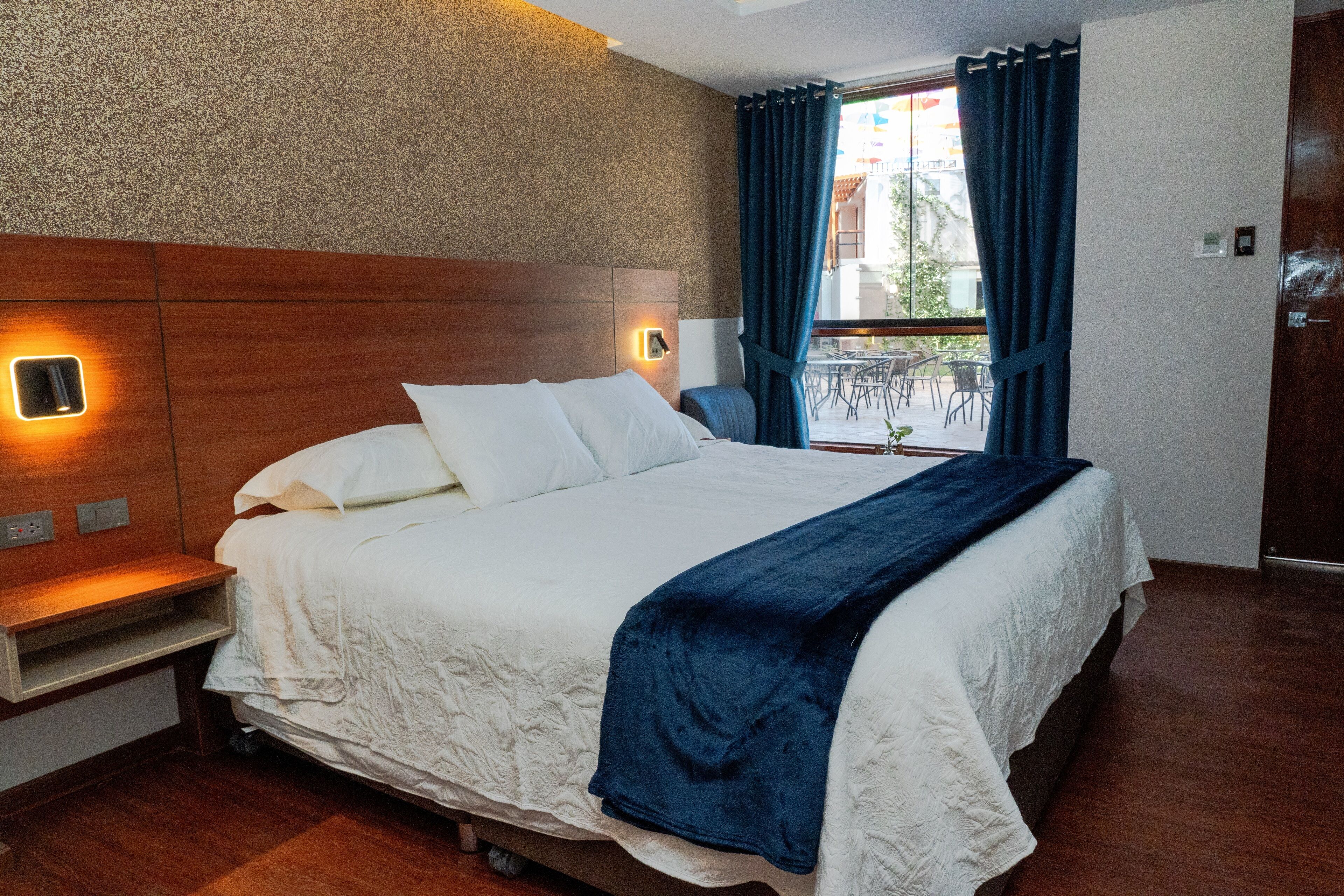 Quarto de hotel com uma cama grande, cabeceira de madeira, mesa de cabeceira e janela com cortinas.