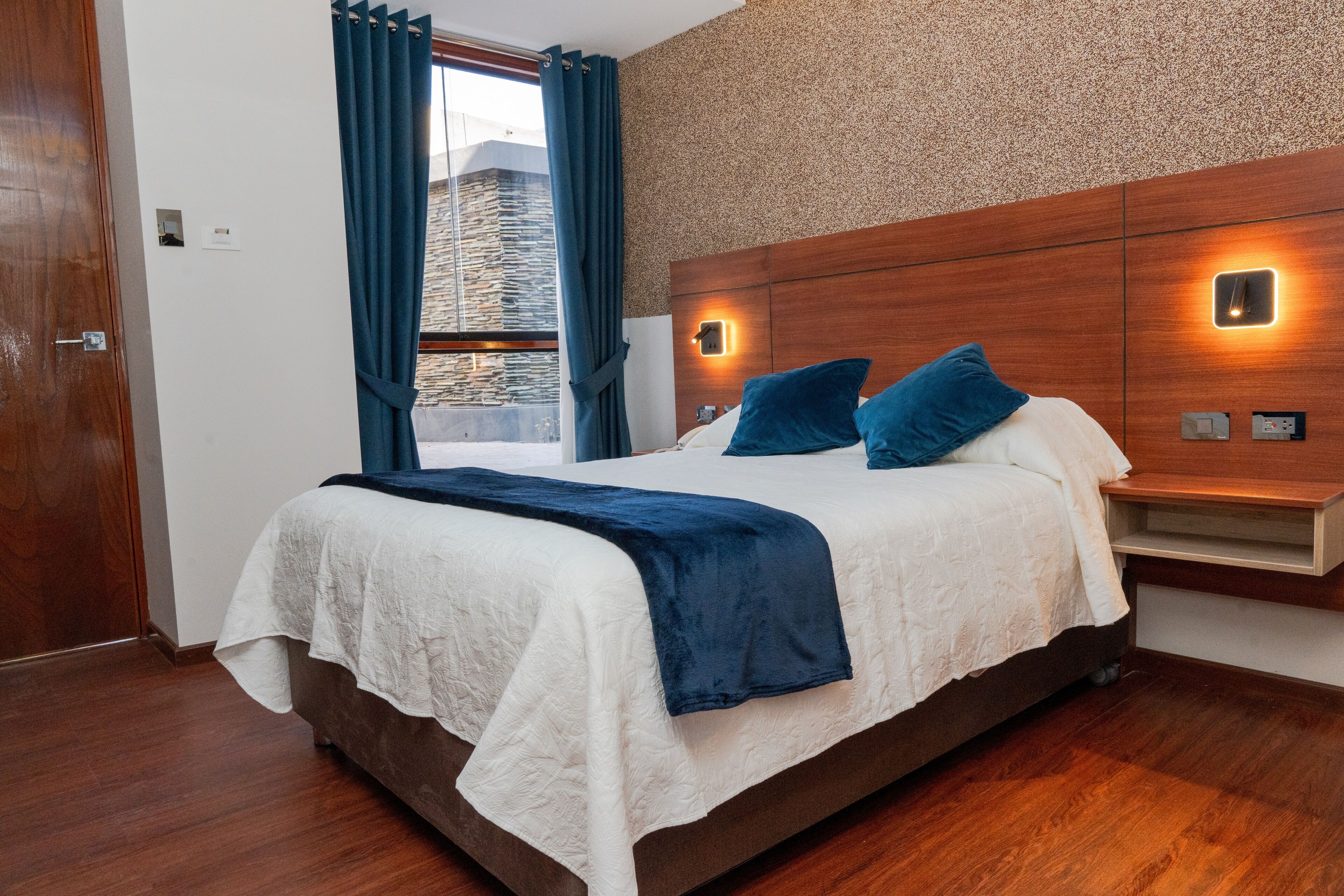 Quarto com cabeceira de madeira, colcha branca, duas almofadas azuis e mesa de cabeceira com luminária.