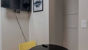 Quarto com uma mesa redonda preta, duas cadeiras amarelas, uma TV de tela plana na parede e duas molduras com quadros.