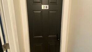 Uma porta de madeira escura com moldura branca, uma pequena placa acima e um chão de madeira que leva a um corredor.