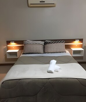 Uma cama bem arrumada com um edredom cinzento, duas almofadas e uma toalha branca enrolada. Há duas mesas de cabeceira com abajures, e um aparelho de ar condicionado fixado na parede acima da cama.