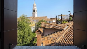 Visão através de uma porta de um edifício com telhado de azulejos, uma igreja com torre de sino e vegetação ao redor.