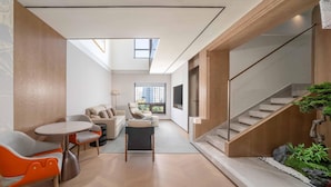Uma sala de estar moderna com um sofá, cadeiras, uma mesa de centro e uma televisão. Há uma escada com guarda-corrimão de madeira e um pequeno jardim interno.