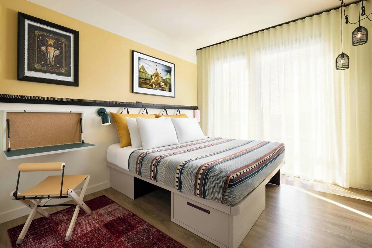 Quarto com cama, uma cadeira pequena, janela com vista e um quadro na parede.