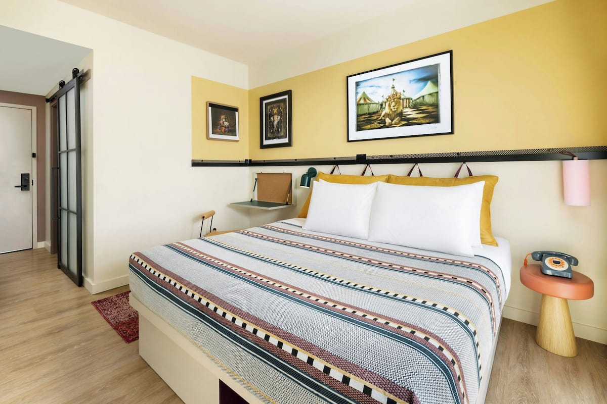 Quarto com cama, mesa de cabeceira com abajur e telefone fixado na parede.
