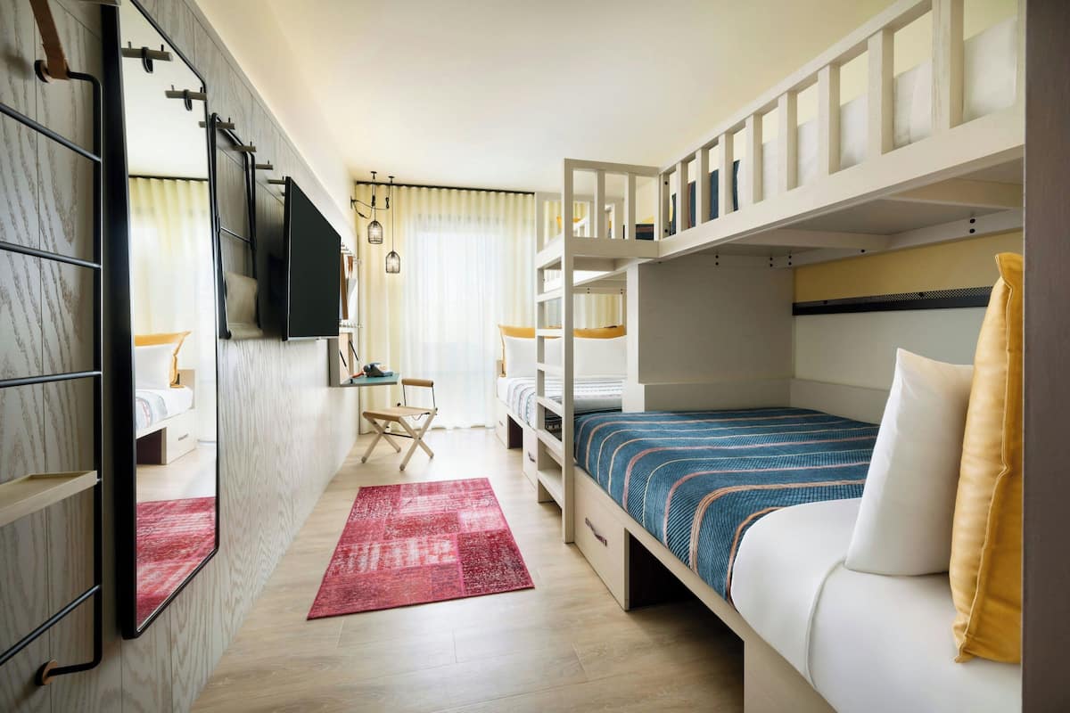 Quarto de hotel compacto com beliche, uma escrivaninha com cadeira, televisão fixada na parede e um tapete vermelho no chão.