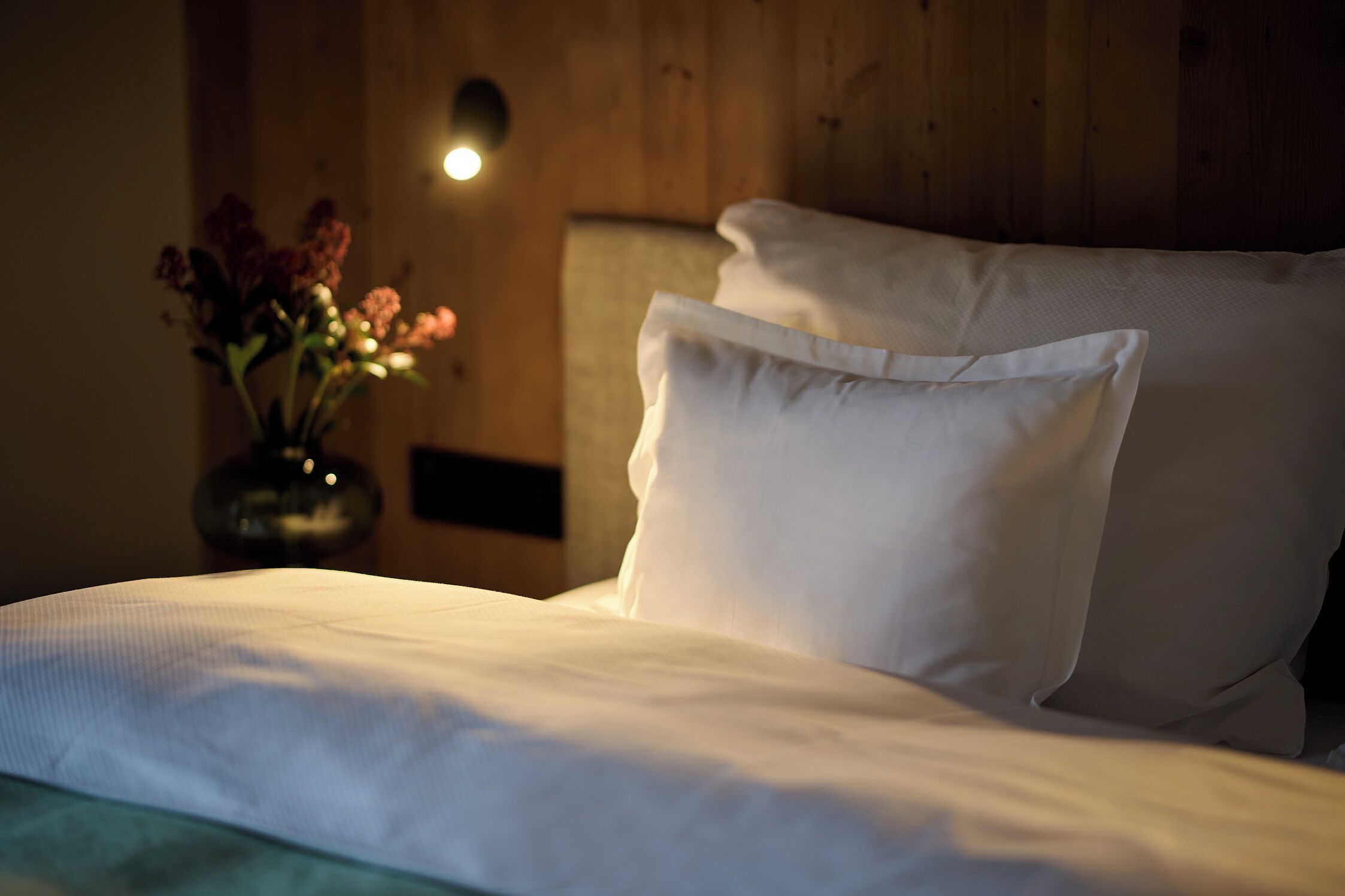 Cama bem arrumada com lençóis e almofadas brancos, um vaso com flores e um luminário fixado na parede.