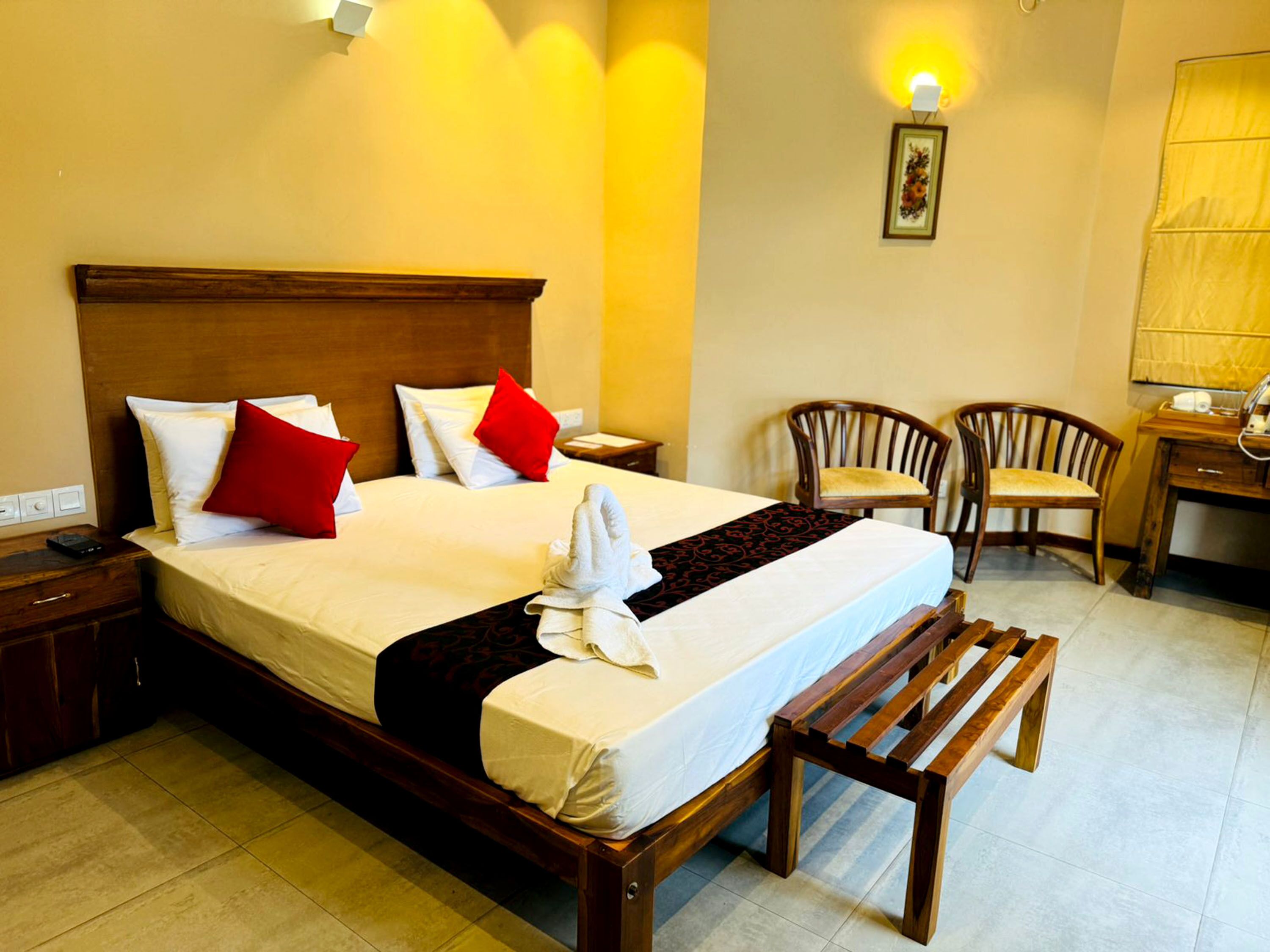 Quarto de hotel com uma cama grande, duas cadeiras de madeira, uma escrivaninha e uma mesinha de cabeceira.