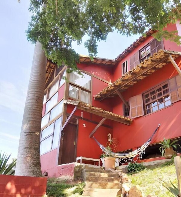 Um edifício vermelho com uma escadaria de madeira que leva a uma porta, cercado por vegetação e uma rede.