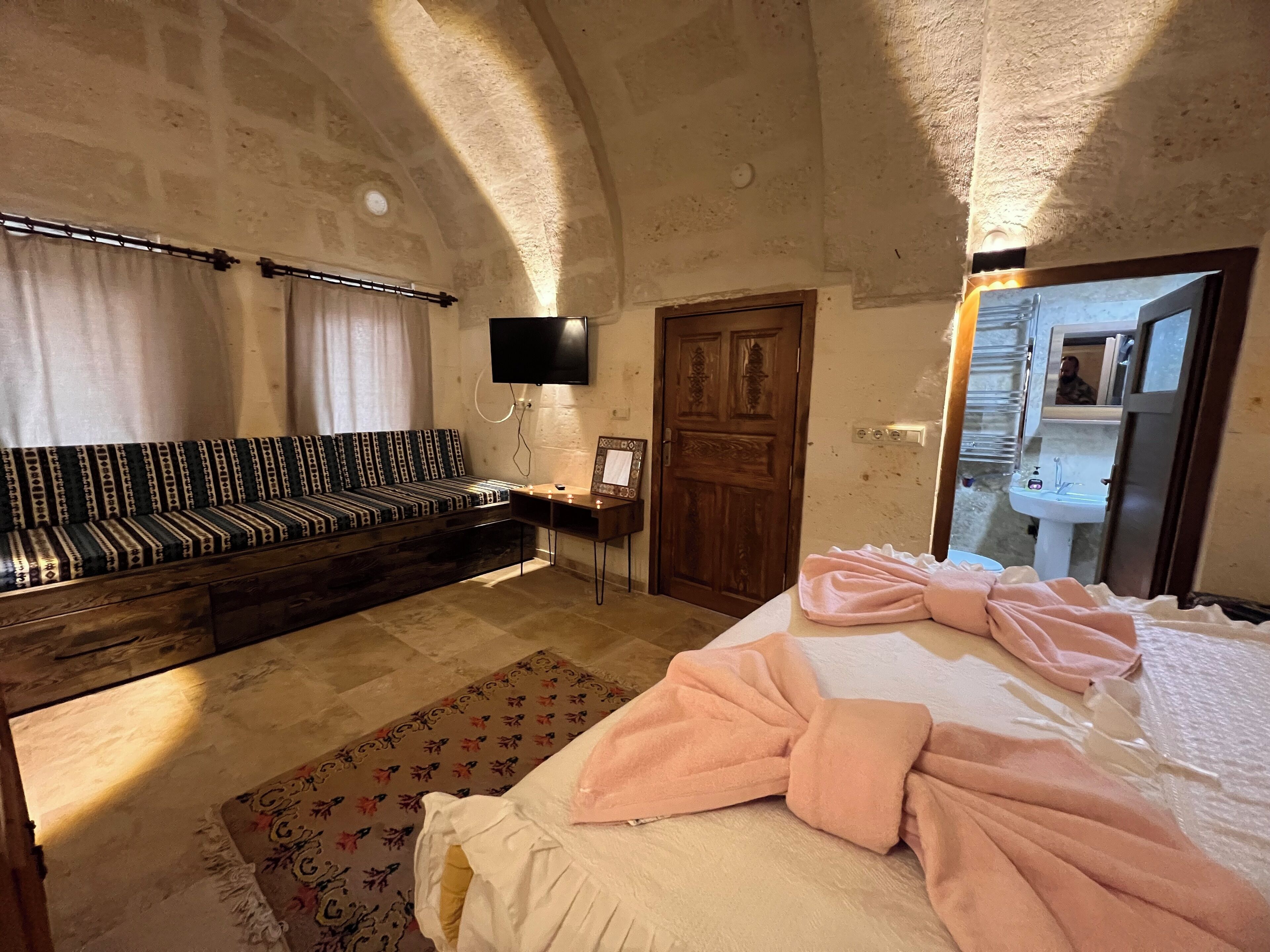 Quarto aconchegante com um banco de madeira, uma cama com toalhas cor-de-rosa, uma televisão e uma porta de madeira.