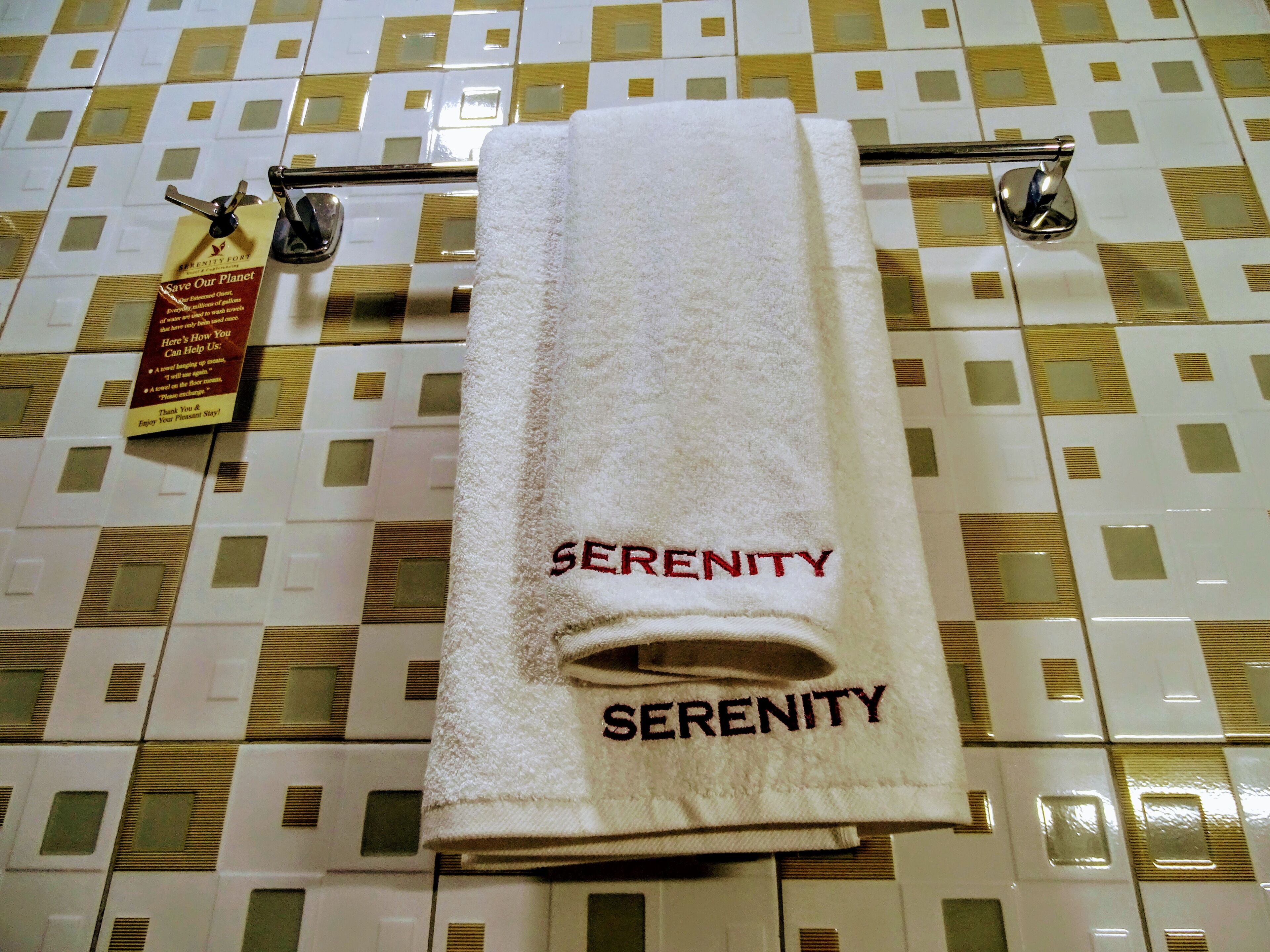 Toalha com a inscrição "Serenity" bordada, pendurada em um porta-toalhas em um quarto com paredes de azulejos estampados.