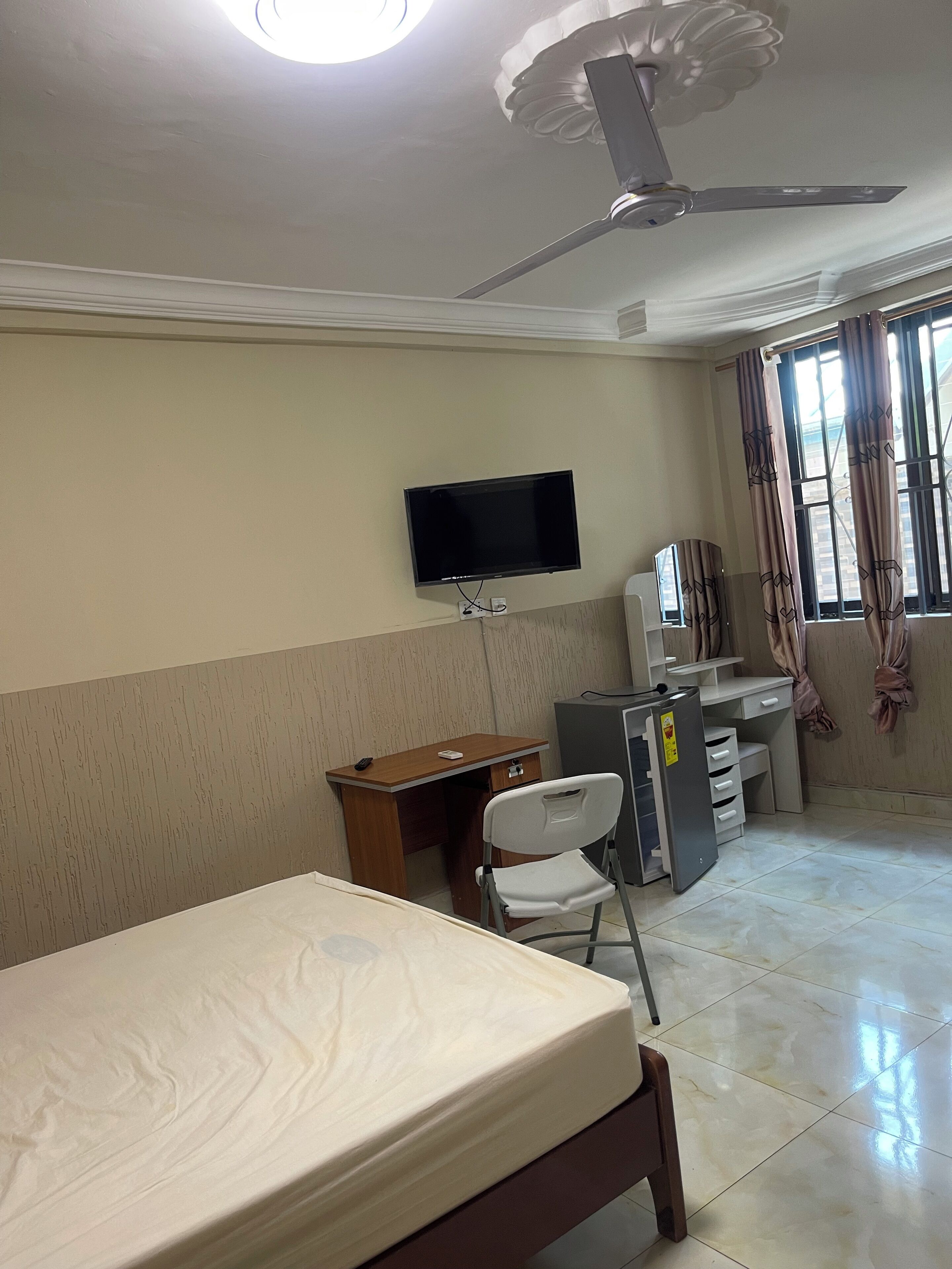 Quarto de hotel com uma cama, uma escrivaninha, uma cadeira, uma TV fixada na parede, um ventilador de teto e uma janela com cortinas.