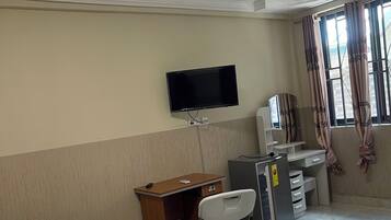 Quarto de hotel com uma cama, uma escrivaninha, uma cadeira, uma TV fixada na parede, um ventilador de teto e uma janela com cortinas.