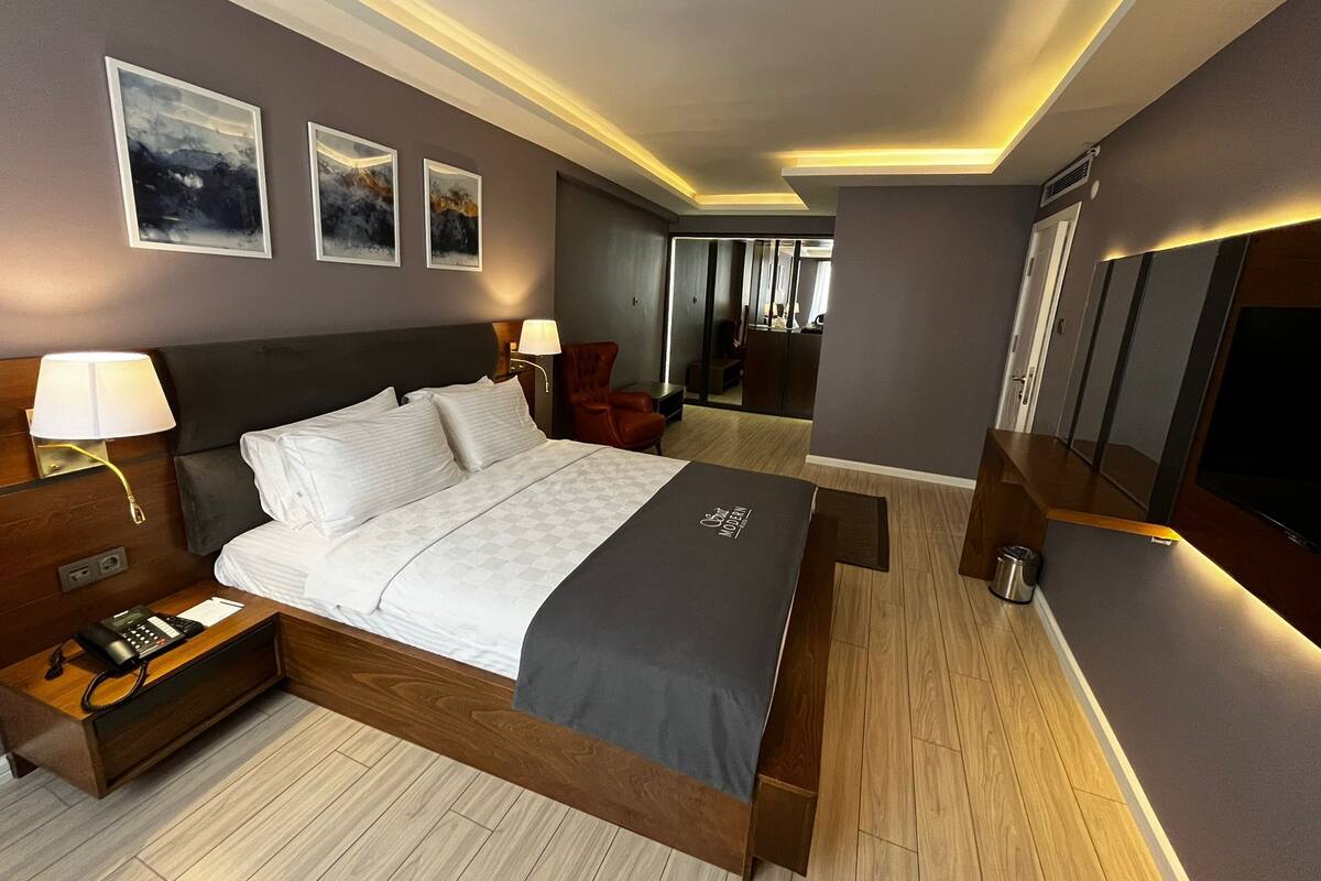 Quarto de hotel moderno com uma cama grande, mesas de cabeceira, escrivaninha, uma cadeira, televisão e obras de arte nas paredes.