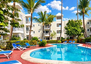 Área de piscina do resort, com piscina azul cristalina, espreguiçadeiras, palmeiras e um edifício com varandas.