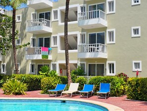Área à beira da piscina com espreguiçadeiras e um edifício com varandas.