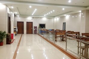 Um hall espaçoso com piso lustroso, bancos de madeira, guarda-corpo de vidro e uma área de recepção com balcão e plantas em vasos.