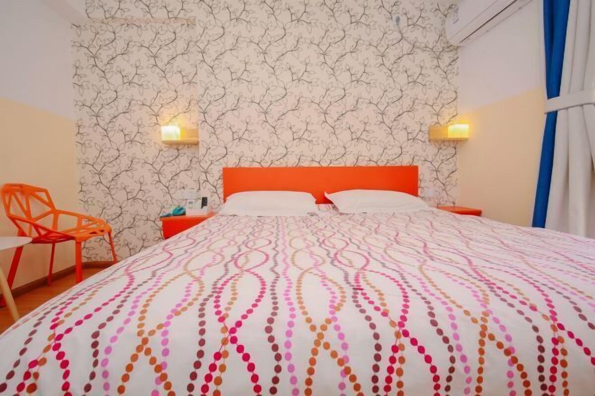 Quarto de hotel com papel de parede estampado, cama com cabeceira laranja, duas mesas de cabeceira, uma cadeira e luminárias fixadas na parede.