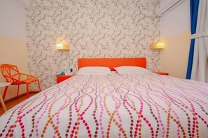 Quarto de hotel com papel de parede estampado, cama com cabeceira laranja, duas mesas de cabeceira, uma cadeira e luminárias fixadas na parede.
