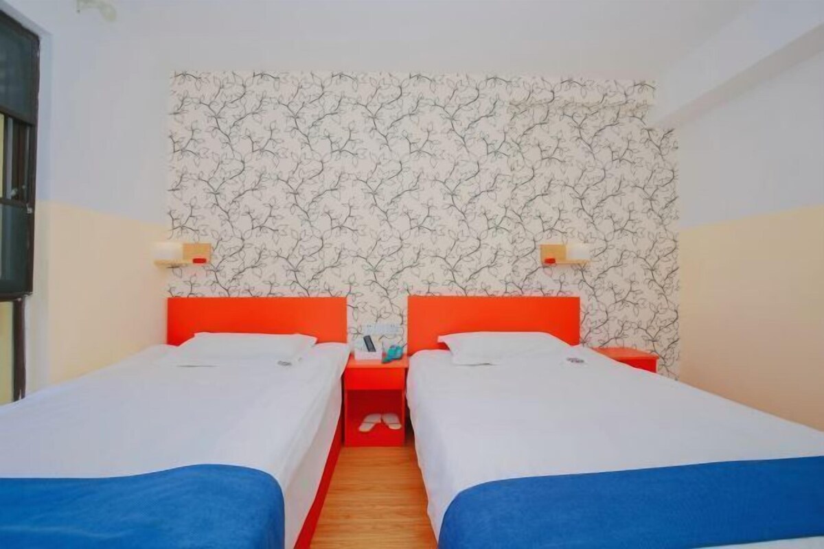 Quarto com duas camas, cada uma com cabeceira vermelha, mesa de cabeceira pequena e parede com padrão de folhas.