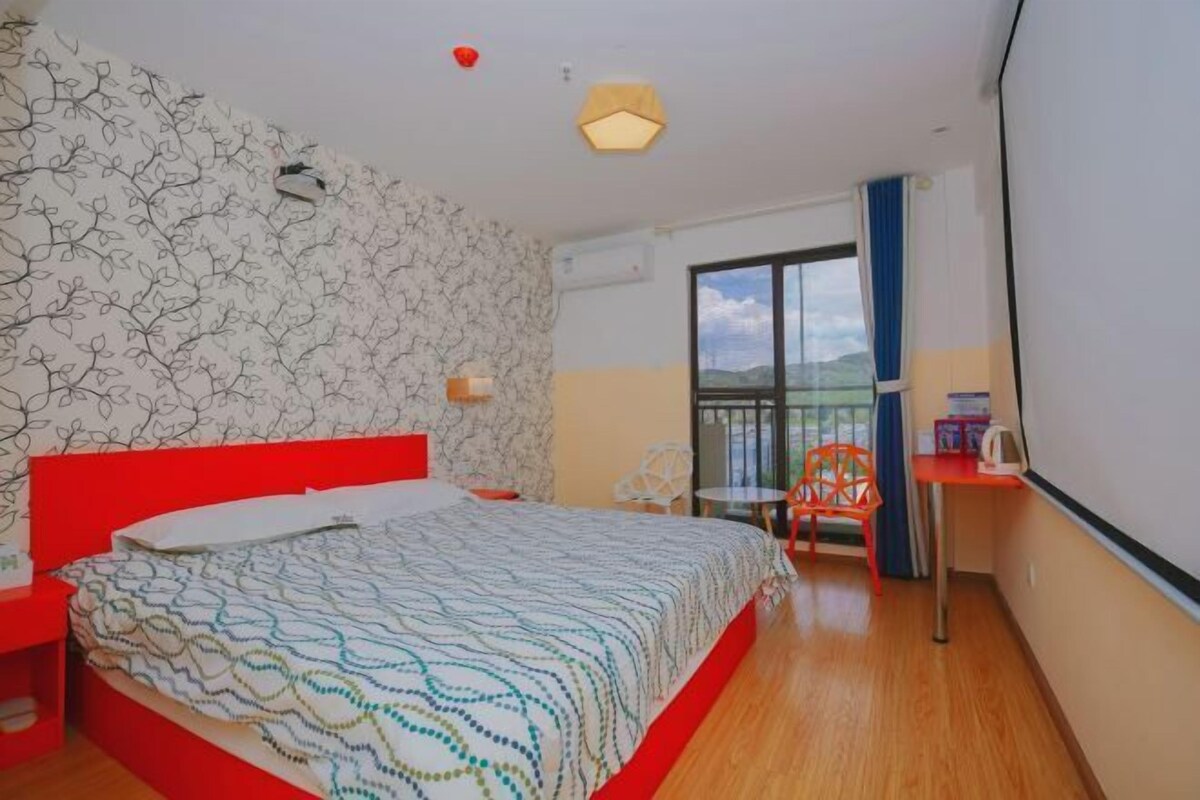 Quarto com cama vermelha, uma mesinha, uma cadeira e janela com vista.