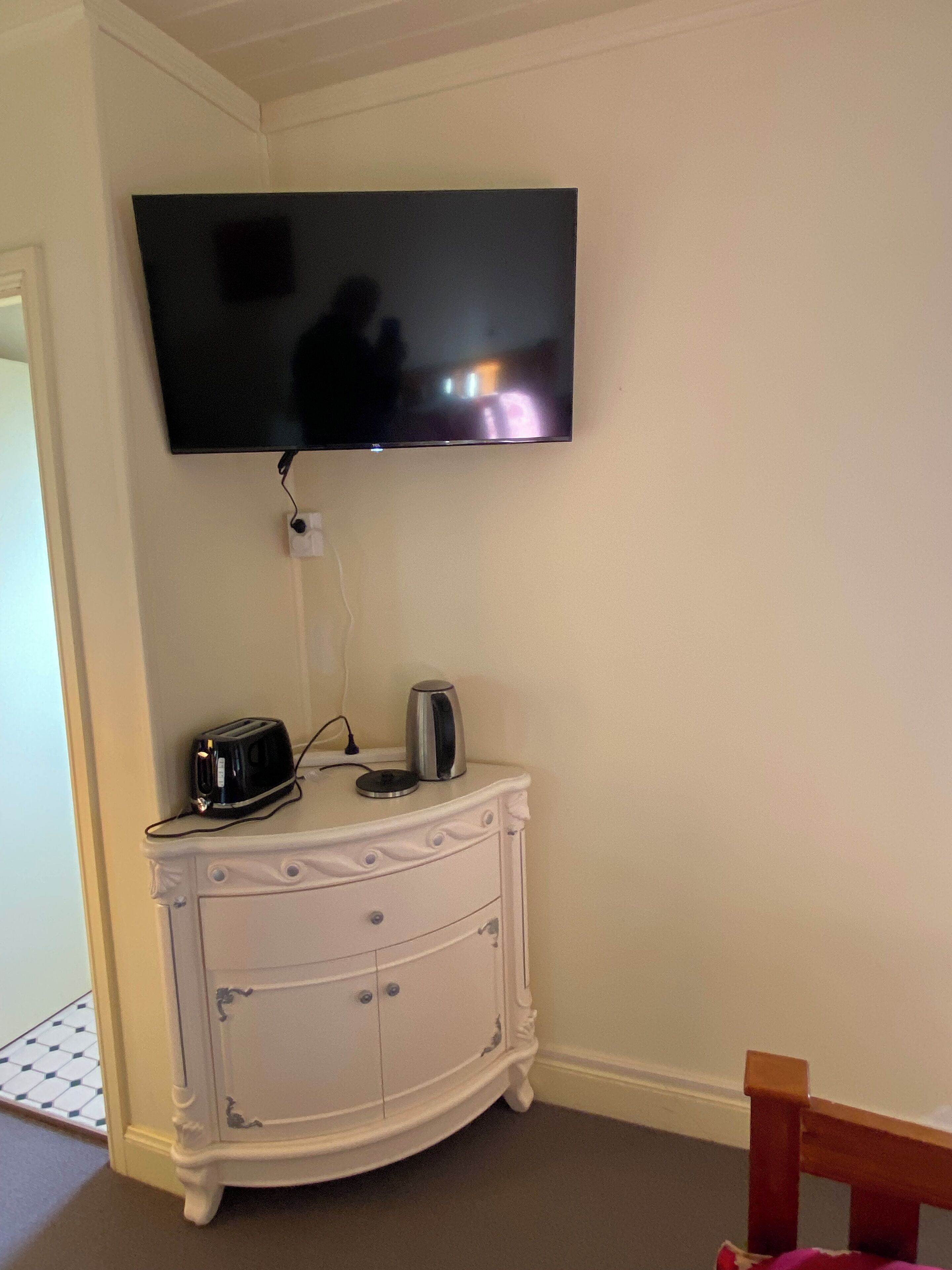 Quarto com televisão de tela plana na parede, armário branco com gavetas e cadeira de madeira.