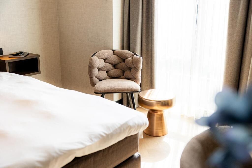 Quarto de hotel com cama, mesa redonda de madeira, poltrona estofada e mesa de cabeceira com abajur.