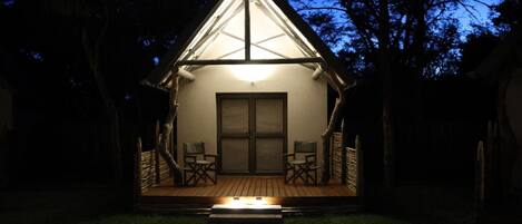 Uma cabana pequena com telhado triangular, deck de madeira e duas cadeiras na parte externa.