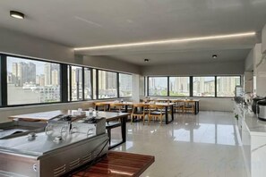 Cozinha moderna com uma ilha grande, mesas de jantar e cadeiras, oferecendo vista para edifícios altos através de amplas janelas.