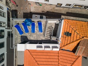 Vista aérea de um terraço com cadeiras de lounge azuis, um edifício branco com telhado de telhas e uma escadaria que leva a um nível inferior.