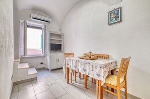 Uma área de refeições com uma mesa posta para dois, uma janela com persiana aberta e um aparelho de ar condicionado branco fixado na parede.