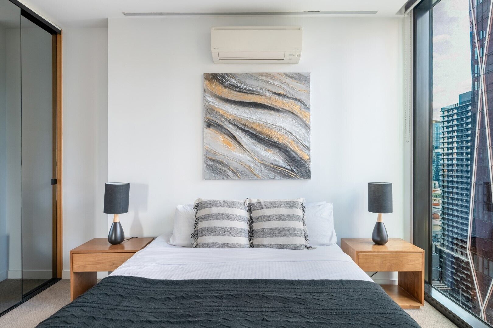 Um quarto moderno com uma cama grande, duas mesas de cabeceira, ar-condicionado na parede e vista para a cidade através de uma grande janela.