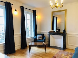 Um quarto com piso de madeira, um sofá preto com almofada decorativa, uma mesa pequena, uma lareira, um espelho grande e cortinas pretas.