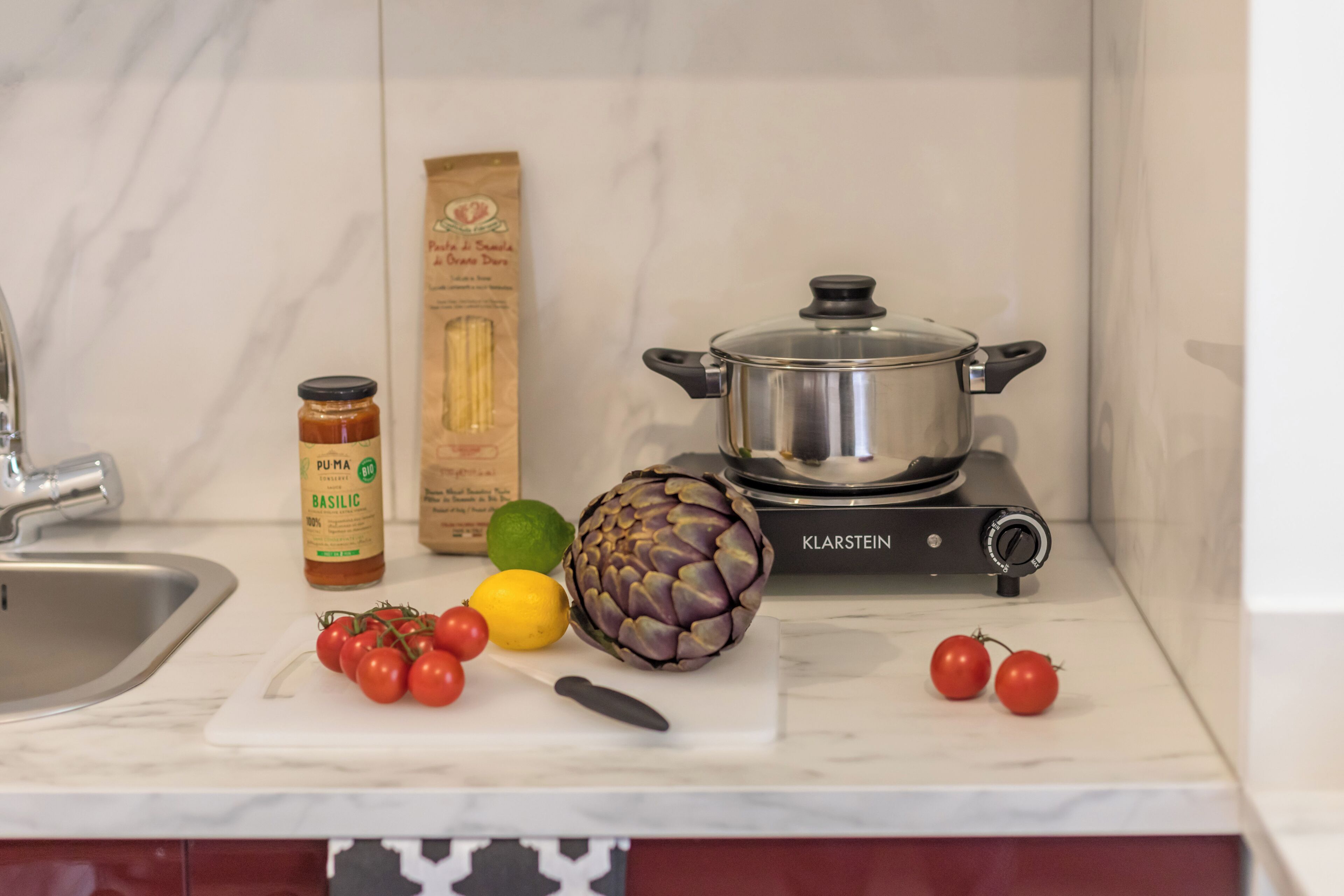 Uma bancada de cozinha com uma panela no fogão, um frasco de molho, tomates, limões e um alcachofra.