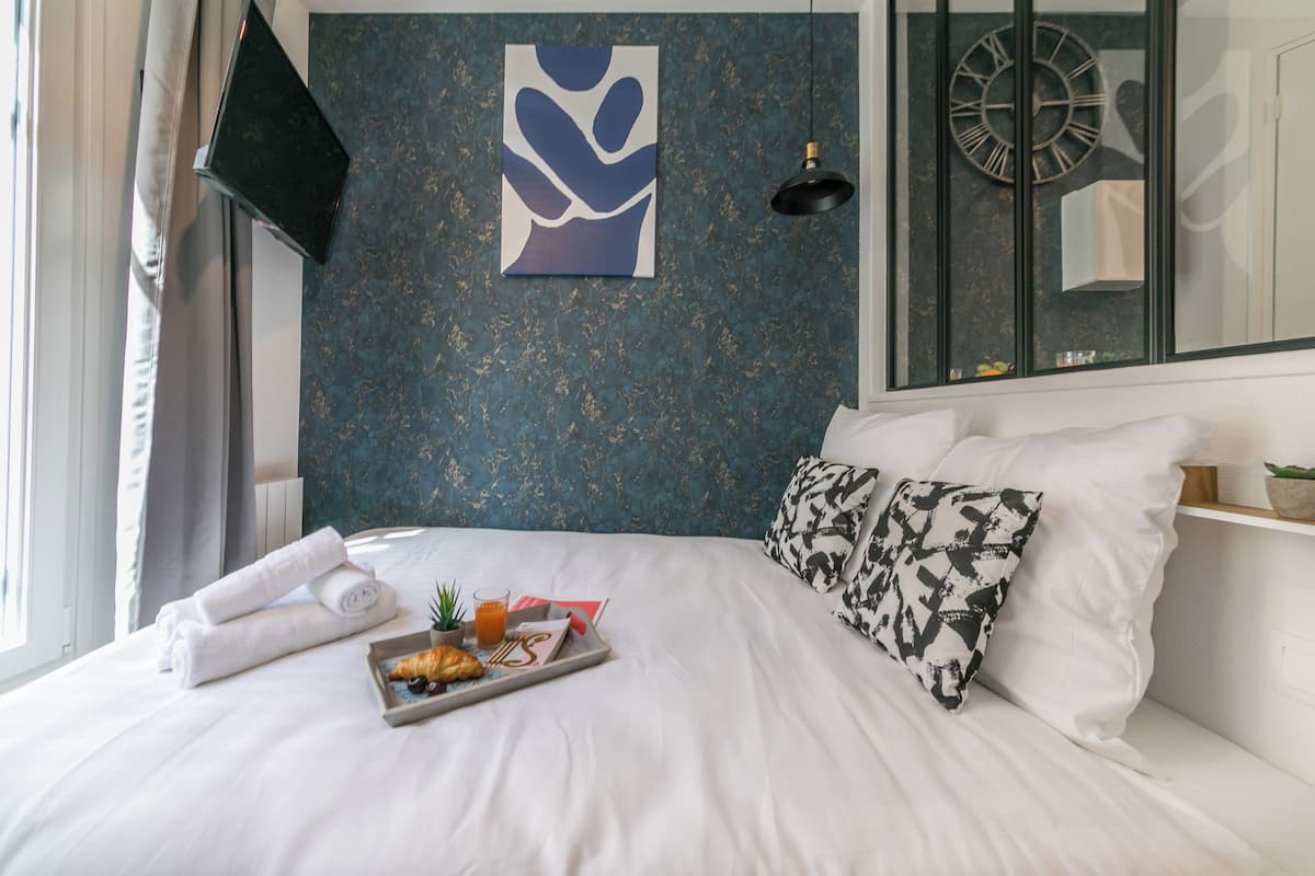 Cama bem arrumada com roupa de cama branca, bandeja com croissants e suco de laranja, e um relógio na parede.