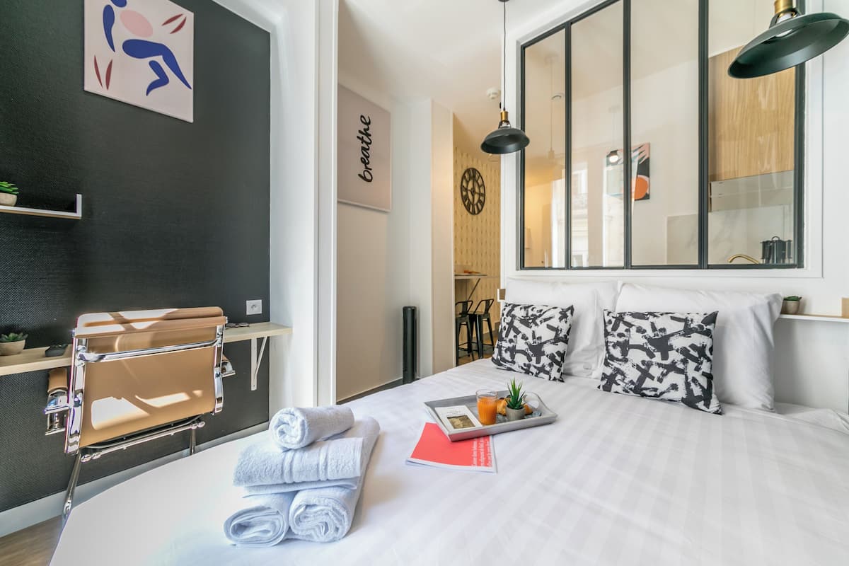 Quarto de hotel moderno com cama, mesa de cabeceira, cadeira, escrivaninha, espelho e um painel decorativo com uma placa.