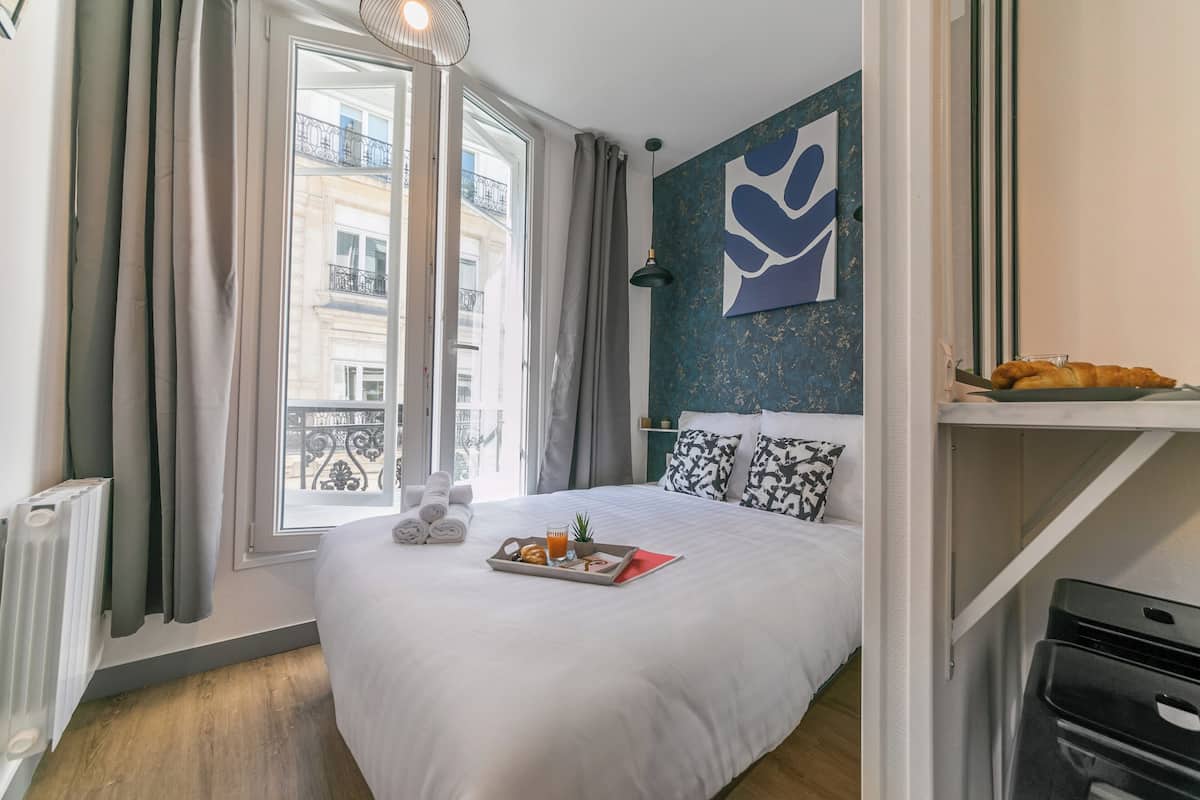 Quarto de hotel com uma cama grande, uma janela com cortinas, um radiador, uma obra de arte na parede e uma bandeja com itens de café da manhã.