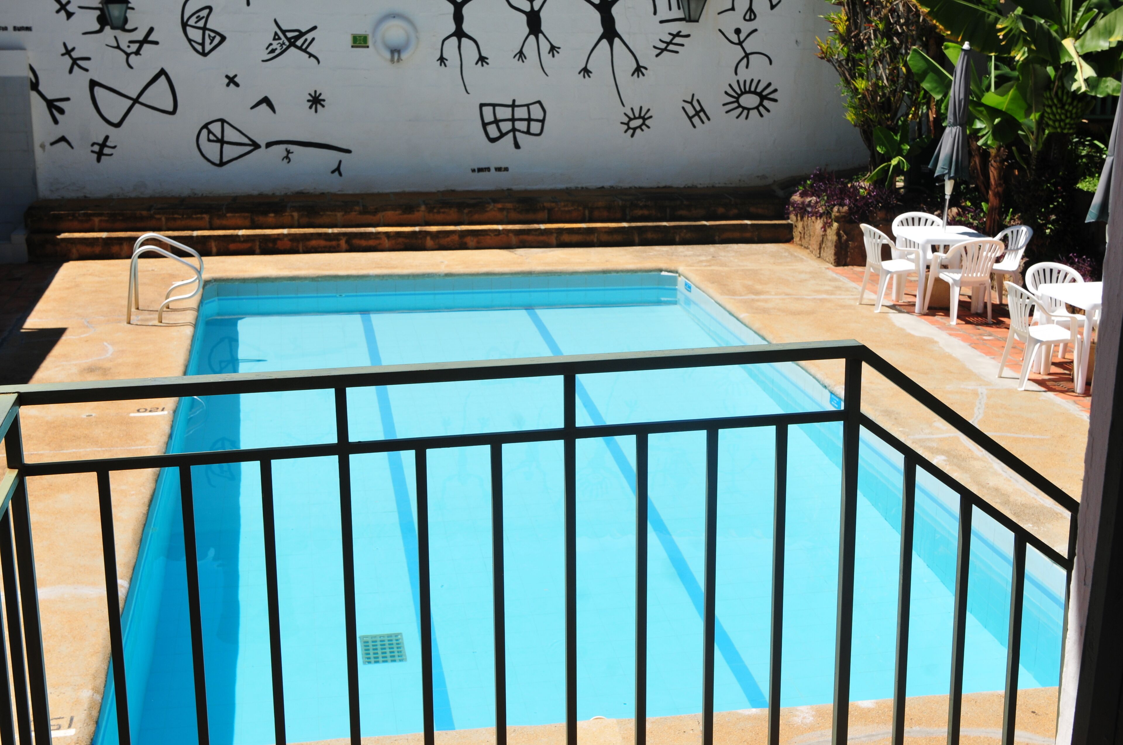 Piscina com grade de metal preta, rodeada por cadeiras e mesas brancas, e uma parede decorada com diversos desenhos pretos.
