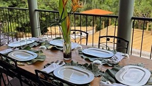 Mesa de jantar posta para seis com pratos, talheres e um vaso de flores, rodeada por cadeiras numa varanda com vista para o verde.