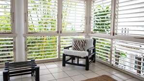 Quarto com cadeira perto de uma janela com persianas brancas, uma mesa pequena e vista para o verde lá fora.