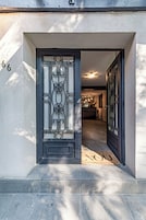 Uma entrada moderna com um portão de metal decorativo, um caminho de pedras e o interior bem iluminado visível pela porta.
