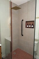 Boxe de vidro com barra de apoio, chão de azulejo e uma prateleira com três garrafas.