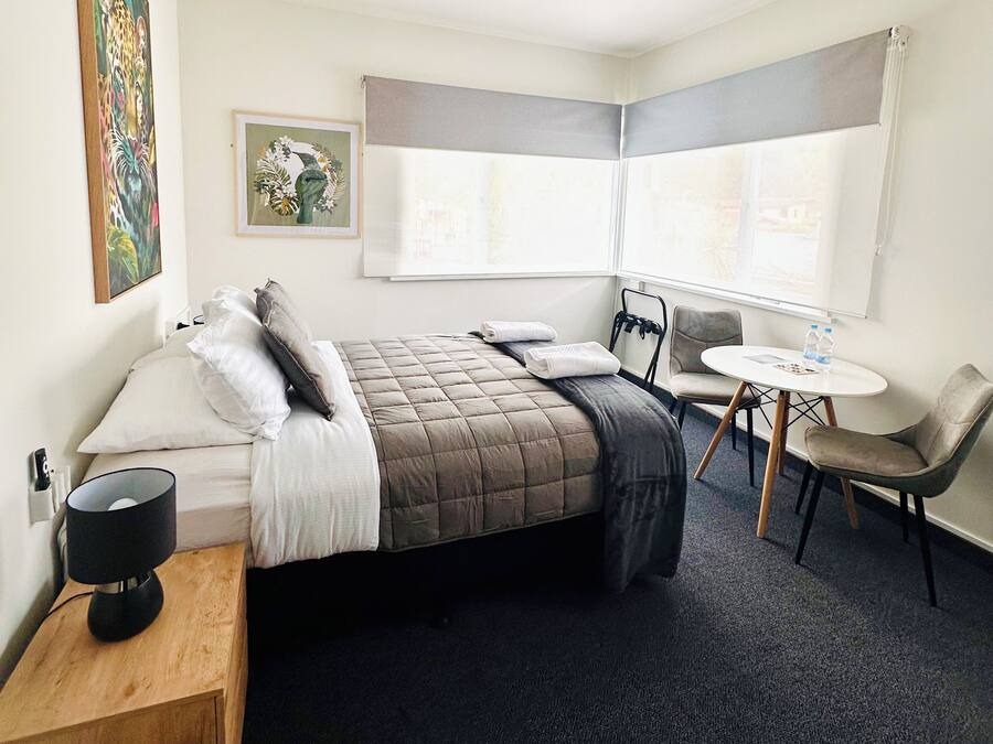 Quarto de hotel com cama, mesa de cabeceira, cadeira, escrivaninha e janela.