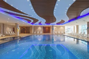 Uma grande piscina coberta com superfície de água azul cristalina, rodeada por espreguiçadeiras e um design de teto moderno.