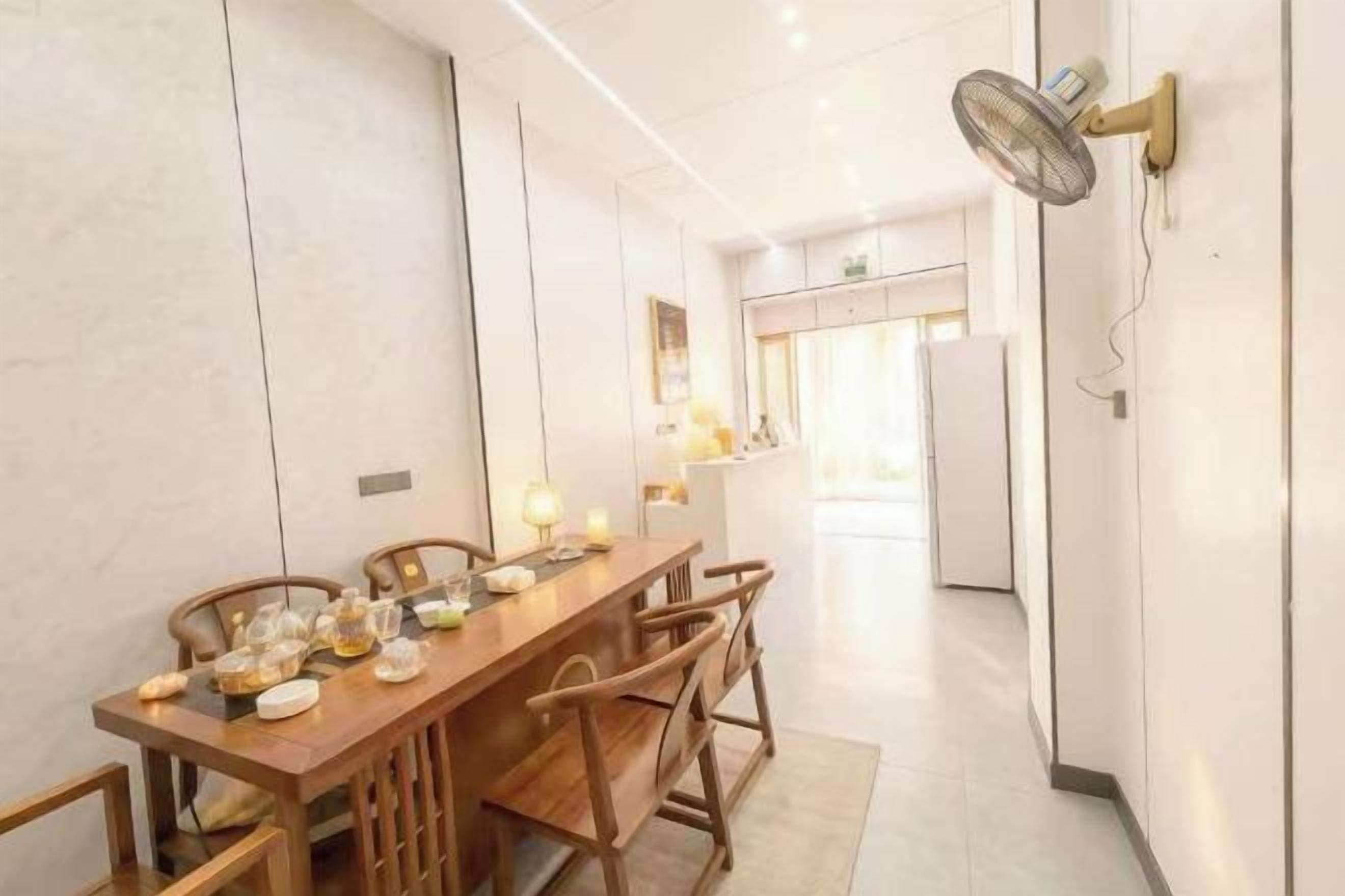 Uma área de refeições com mesa e cadeiras de madeira, um ventilador fixado na parede e uma cozinha ao fundo.