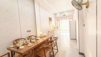 Uma área de refeições com mesa e cadeiras de madeira, um ventilador fixado na parede e uma cozinha ao fundo.